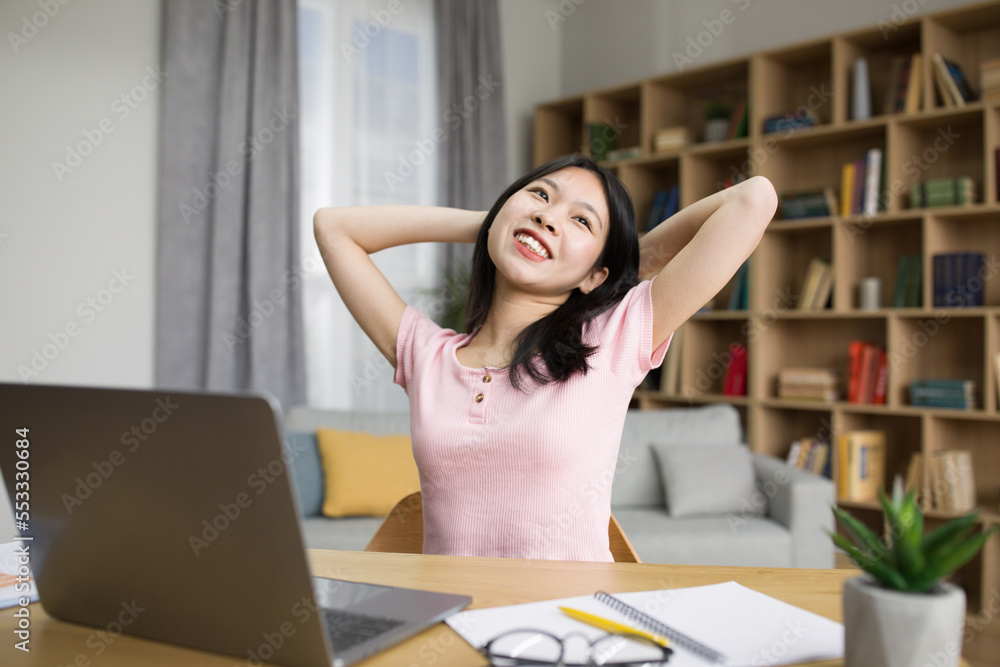 Relaxed asian female student sitting with hands behind head near laptop ...