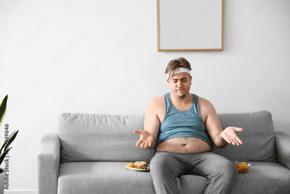 Upset young overweight man with unhealthy food sitting on sofa at home ...