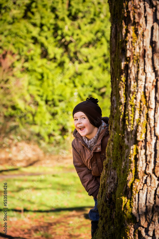 A young boy peeking out from behind a tree with a big smile on his face ...
