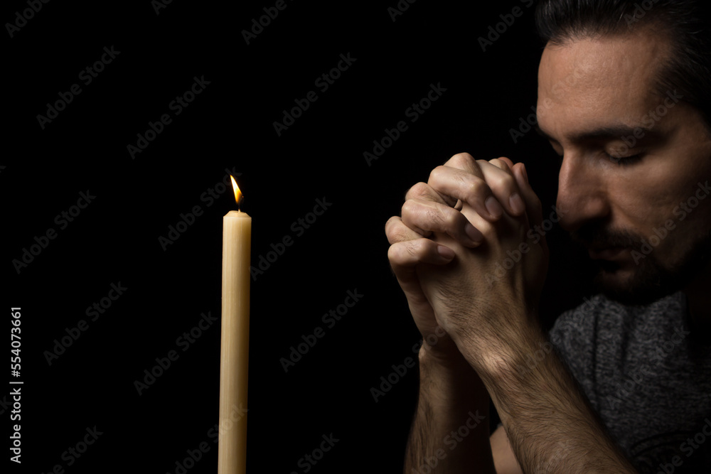 A man folded his hands in prayer next to a burning candle on a black ...