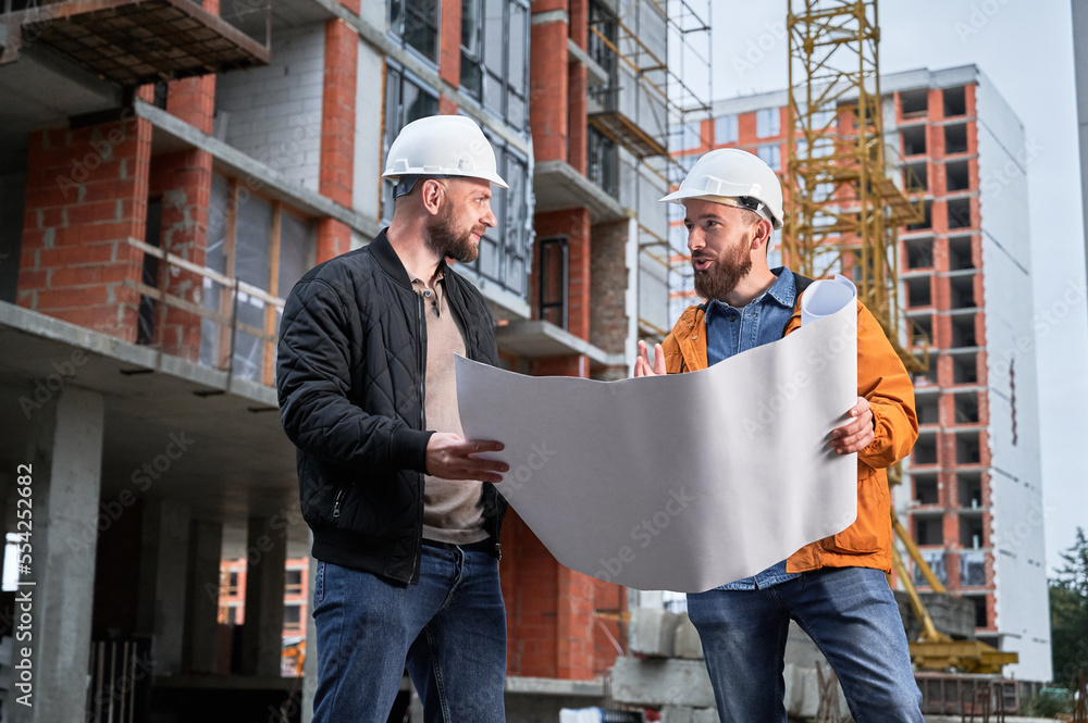 Bearded man in safety helmet holding architectural plan and talking ...