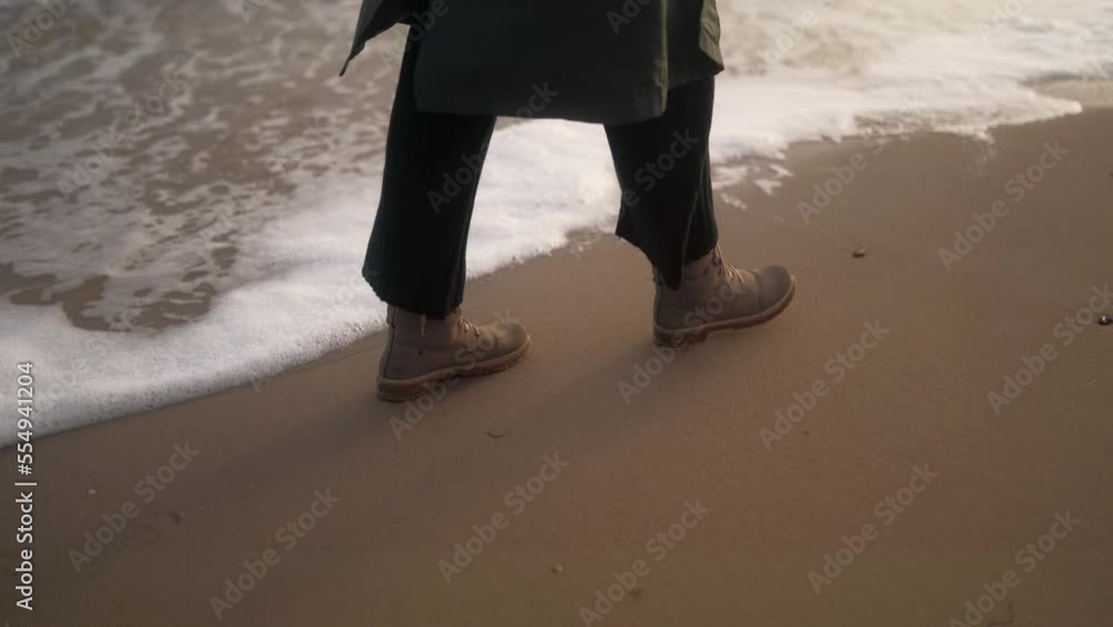 Side view of female feet in boots walking along sea waves foam on sandy ...