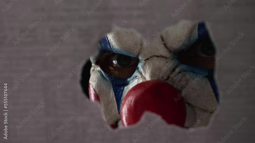A scary clown looks into the camera through a hole in the wall. Close ...