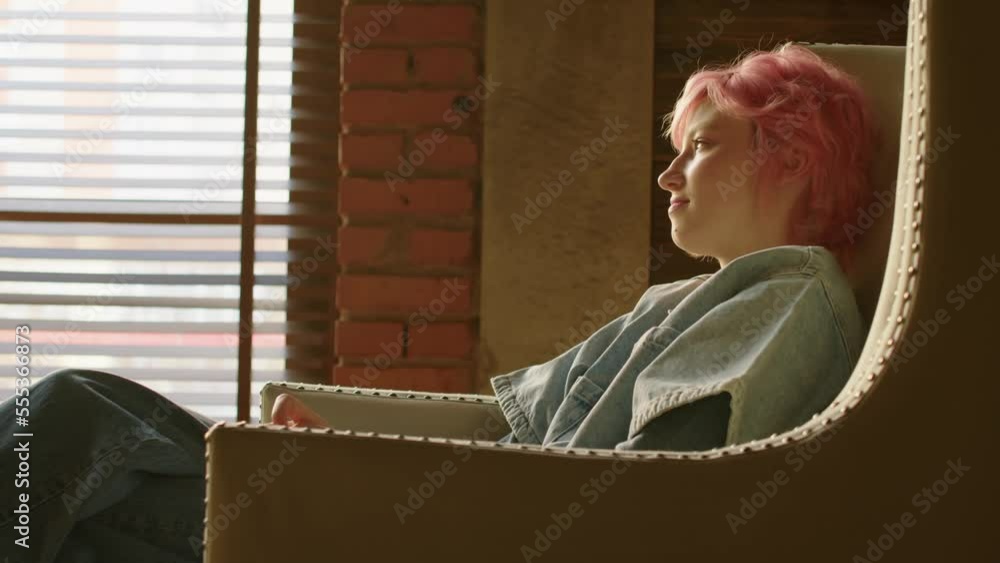 Side view of young gen Z woman with short pink hair sitting in armchair ...