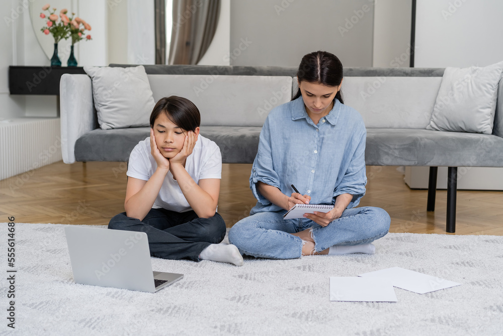 Preteen asian boy looking at laptop while mother writing on notebook at ...