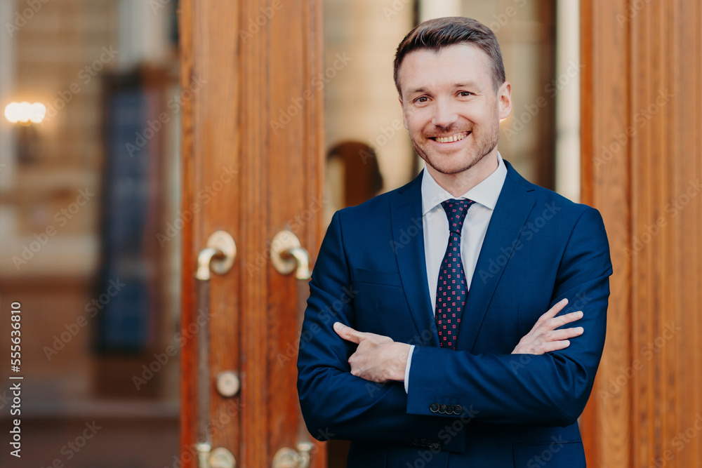 Pleased attractive businessman with positive smile, keeps hands crossed ...