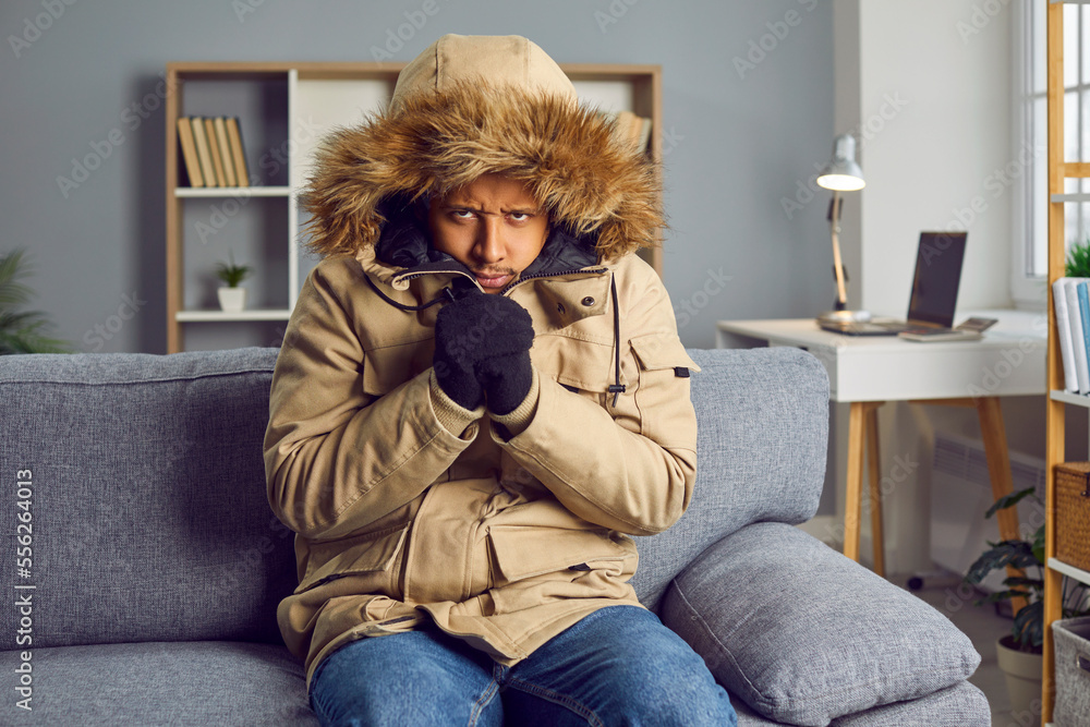 Young man freezing indoors without central heating. African American man wearing warm winter ...