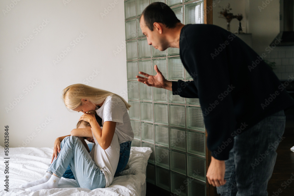 Back view of scared mother and daughter hugging each other sitting on sofa while aggressive ...