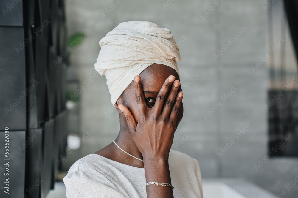 Shy African woman in turban covers face by hand looks at camera through ...