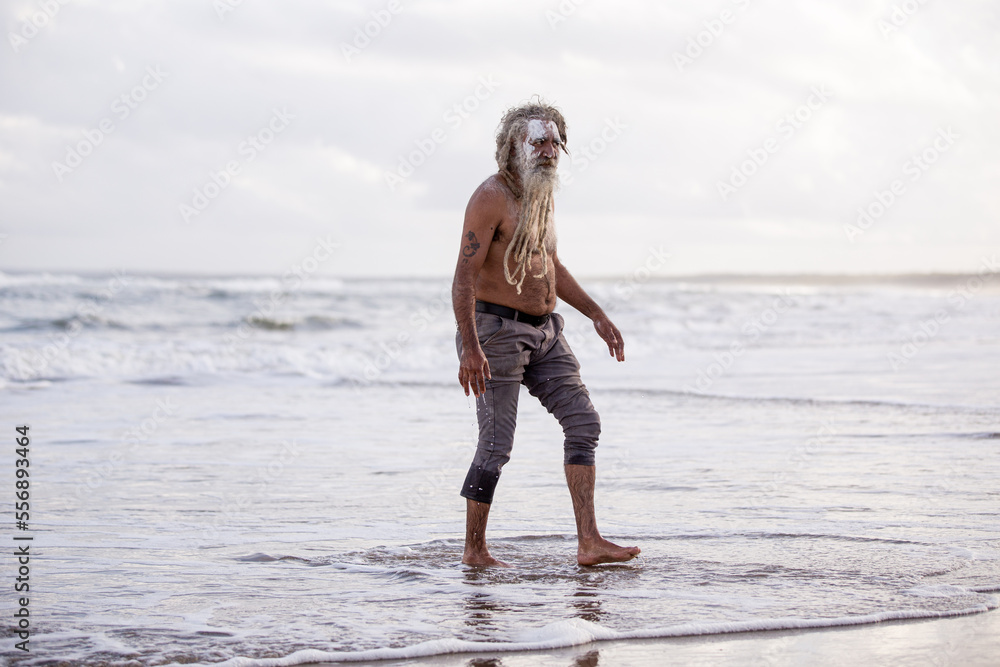 Aboriginal middle aged man walking through water on a beach looking at ...
