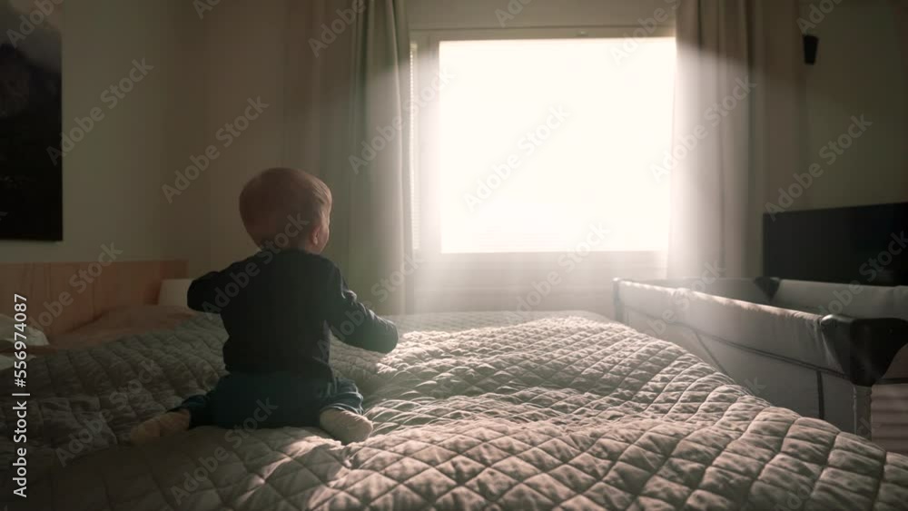 Active blond toddler jumps on soft bed looking out of window ...