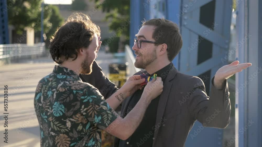 A gay man's rainbow bowtie is fixed by his partner on a blue pedestrian ...