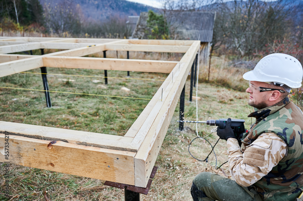 Man building wooden frame house on pile foundation. Worker drilling ...