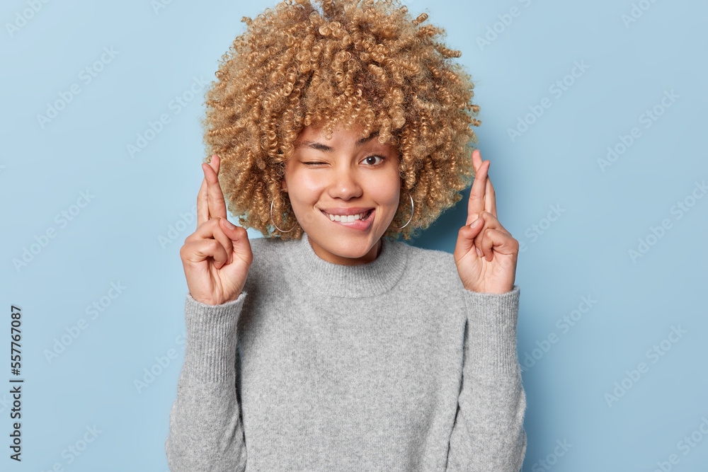 Hopeful positive woman with curly hair bites lips winks eye and crossed ...