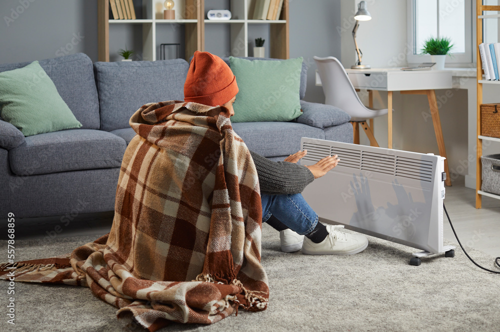 Young woman feeling cold at home, wearing a blanket and a winter hat ...