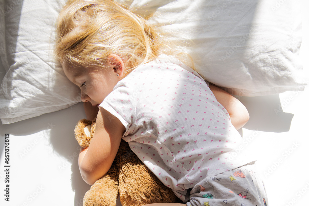 Little Girl Lies Hugging A Soft Toy Bear On A White Bed Top View Flat