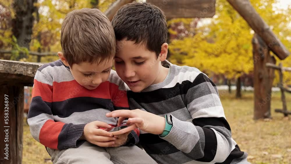 Two little boys playing with mobile phone sitting on the bench outdoors ...