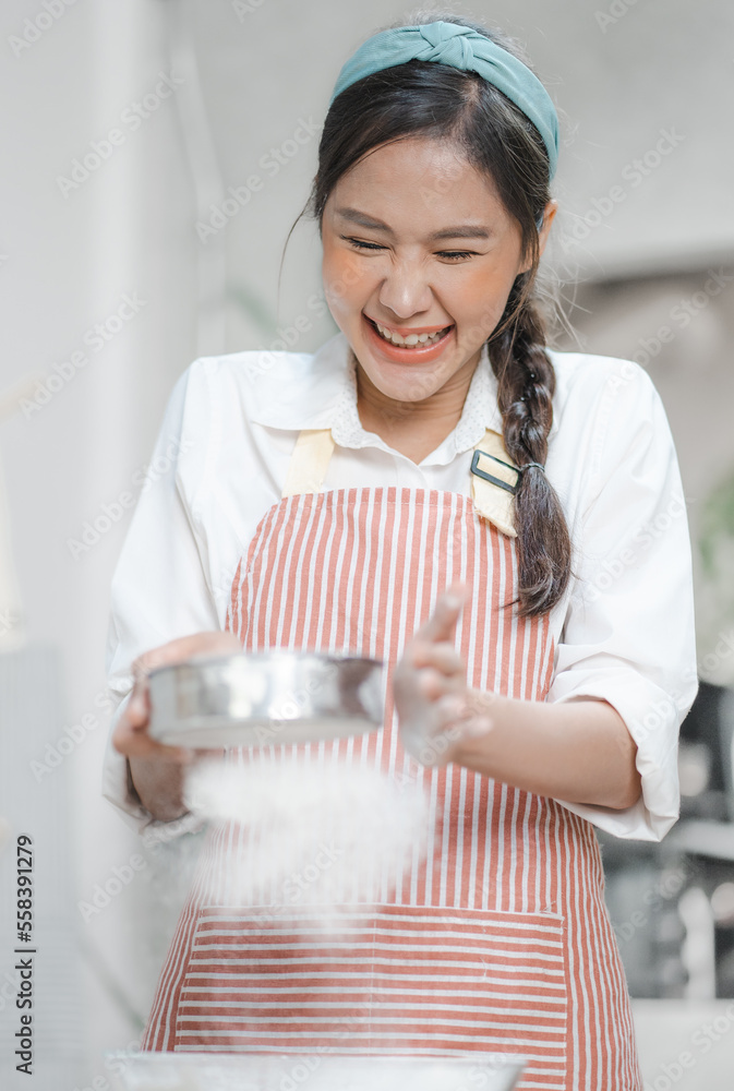 Young woman standing in kitchen sifting flour for homemade bakery or ...