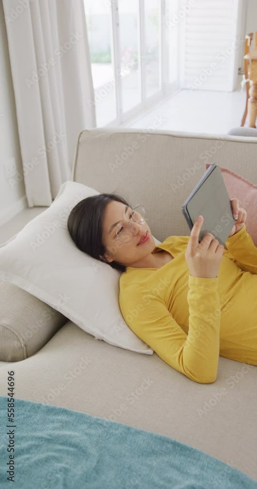 Vertical video of happy asian woman lying on coach and using tablet ...