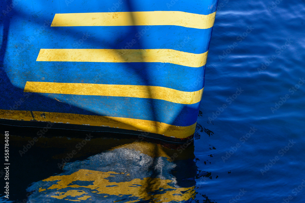 The bow of a large blue ship with yellow stripes reflected in the calm ...