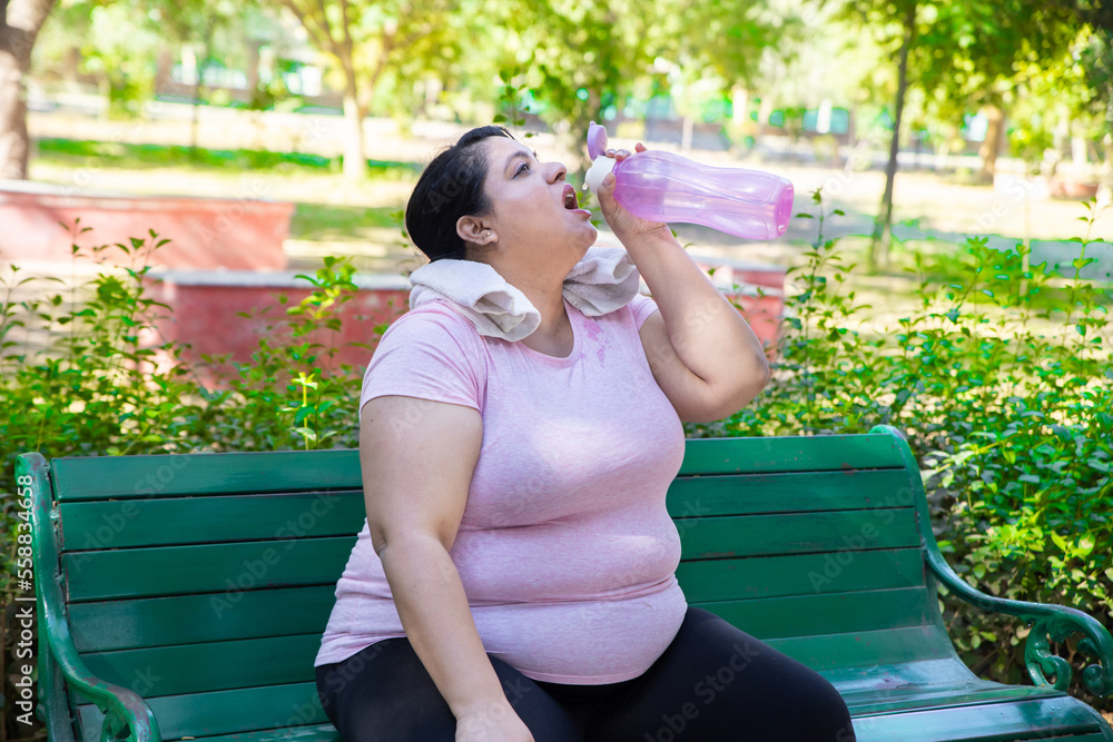 Overweight tired exhausted indian woman sitting drinking water outdoors ...