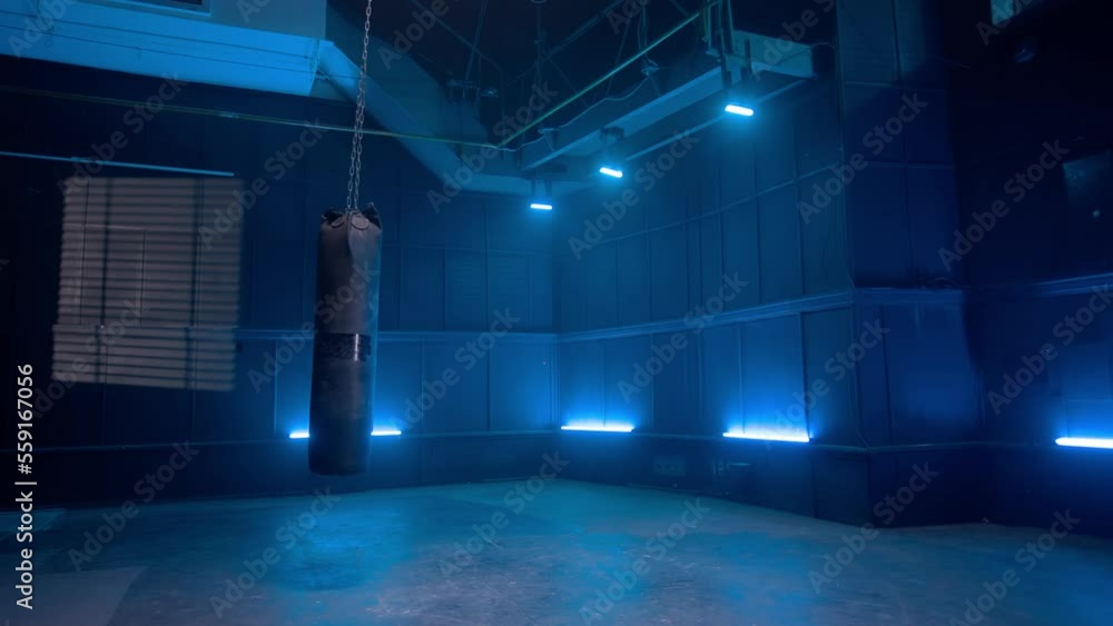 Long black punchbag hanging and swinging from chain in dark empty gym ...