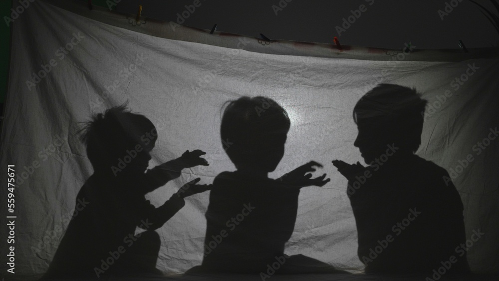 Three children playing shadows on sheet tent lit by lamp in their room ...