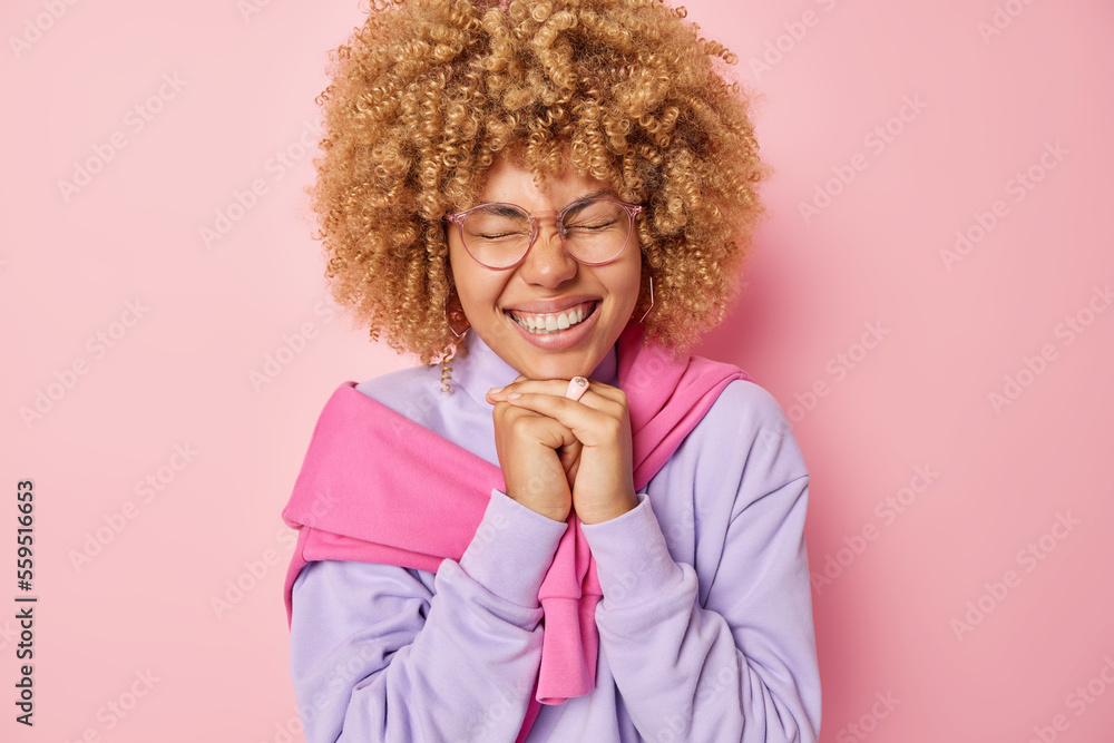 Overjoyed curly haired female model keeps hands under chin laughs ...