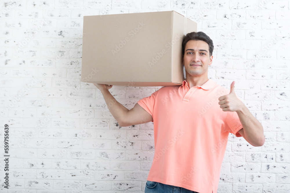 Handsome man carrying huge and heavy box in front of white brick wall ...