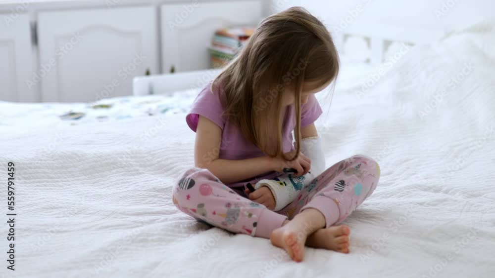 Adorable preschooler girl with a broken arm at home on the bed draws ...