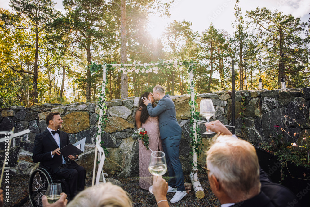 Newlywed couple kissing in front of guests raising toasts and minister ...