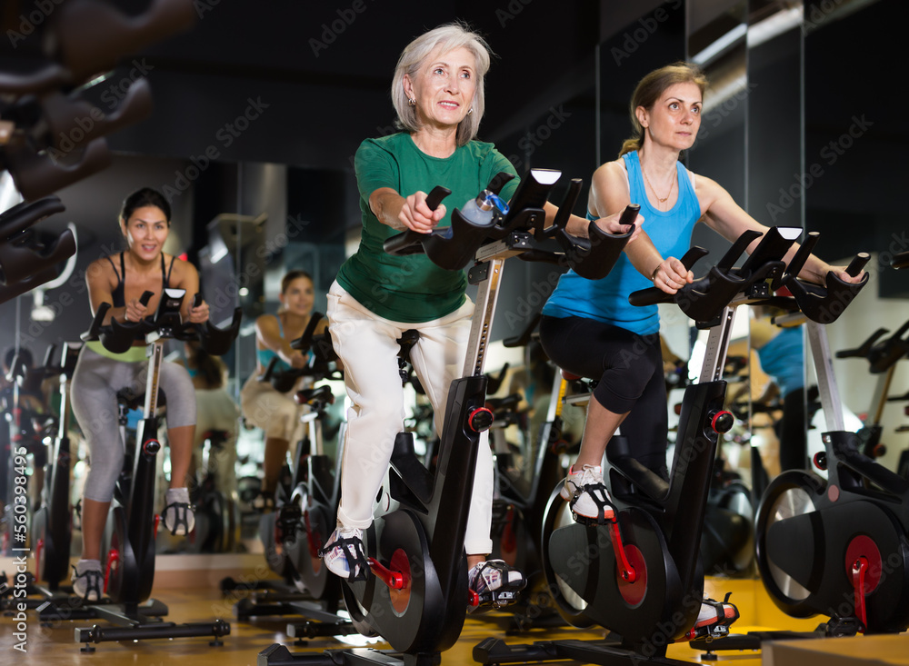 Two positive elderly female riding exercise bike during cycling class ...