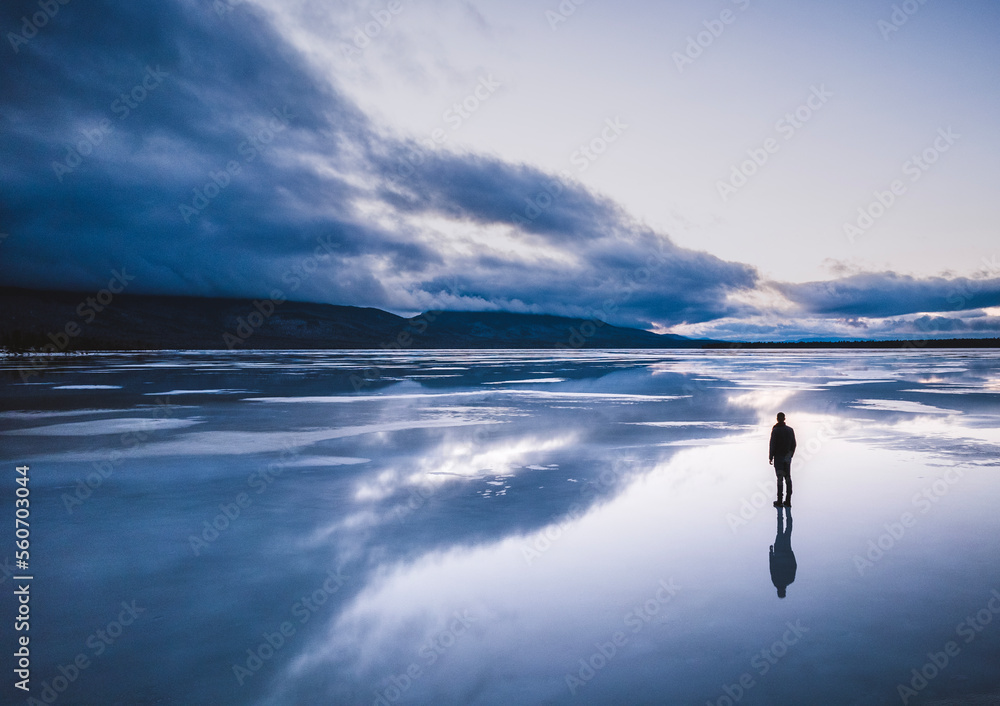 Lone figure stands on frozen lake with reflection and storm clouds ...