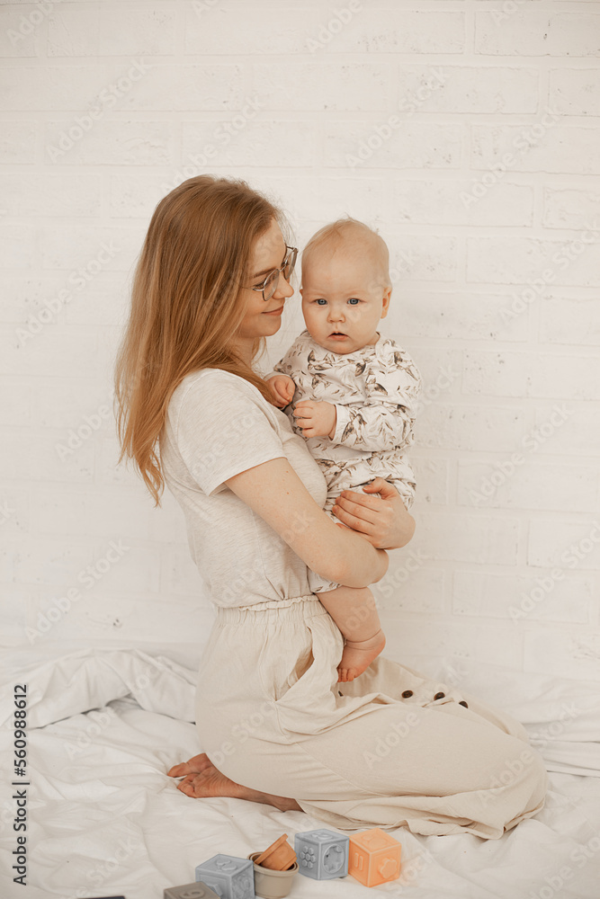 Joyful young mother sit on lap on bed and hold little baby on white ...