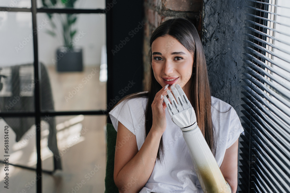 Happy brunette young woman uses artificial bionic hand stands at window ...