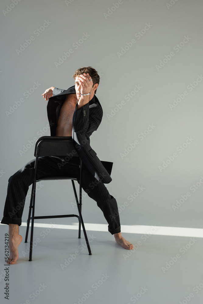 full length of trendy barefoot man in black suit sitting on chair and ...