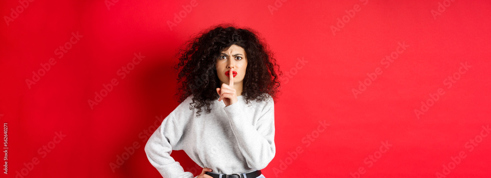 Angry young woman with curly hair and red lips, frowning and shushing ...