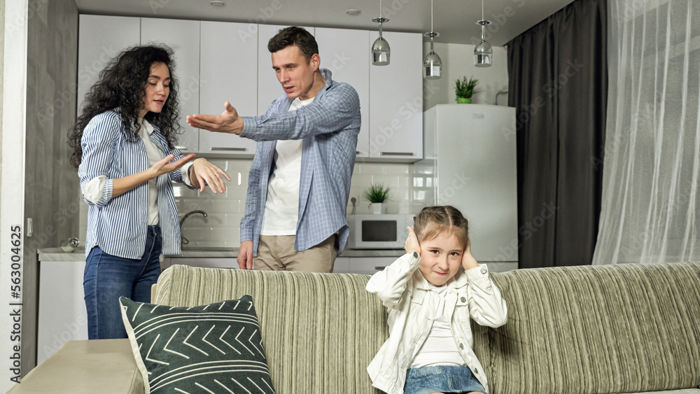 Junior schoolgirl sits closing ears with hands against parents arguing ...