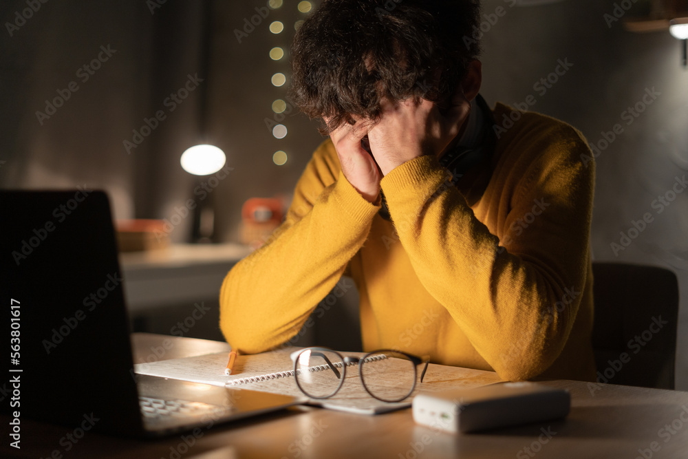Tired and exhausted young man hides eyes with hands at laptop pc late ...