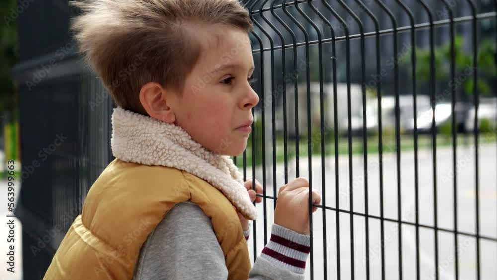 Portrait of lonely boy holding hands on metal fence and looking through ...