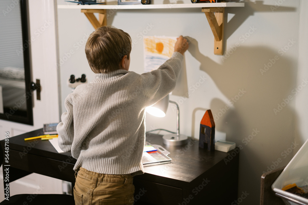 Little boy in his room at his desk holding his drawing to his wall ...