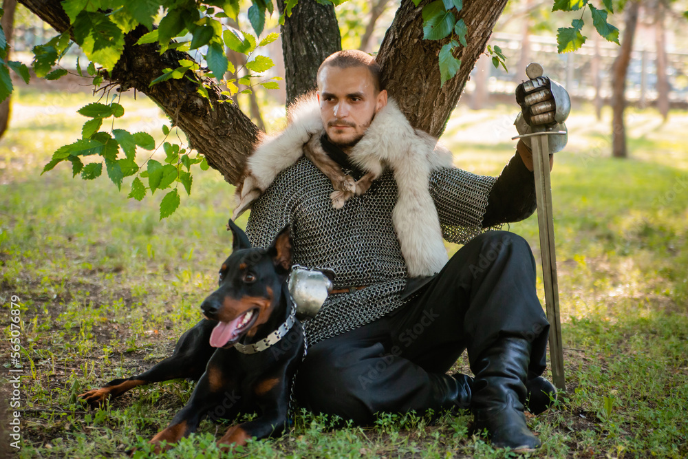 A young man in chain mail with a sword in the forest sits under a tree ...