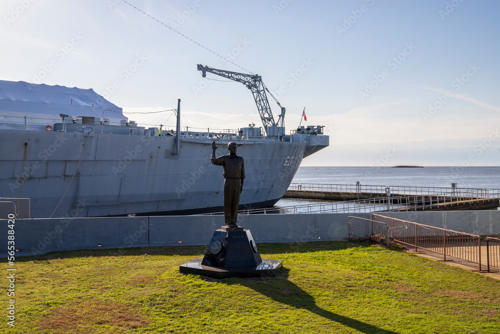 A bronze statue of a man holding up his right hand in front of the USS ...