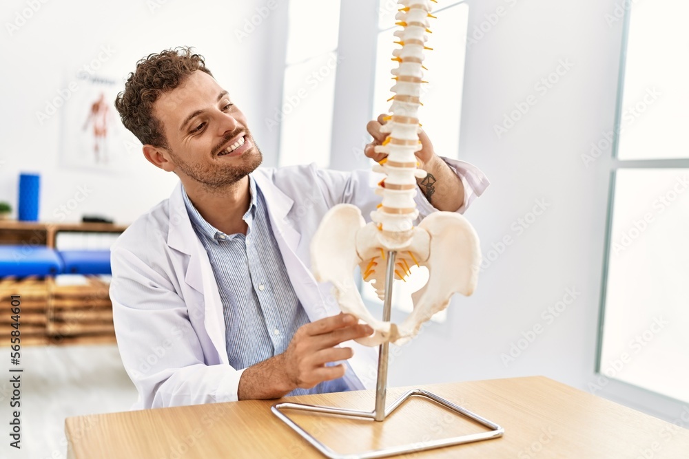 Young hispanic man wearing physiotherapist uniform touching anatomical ...