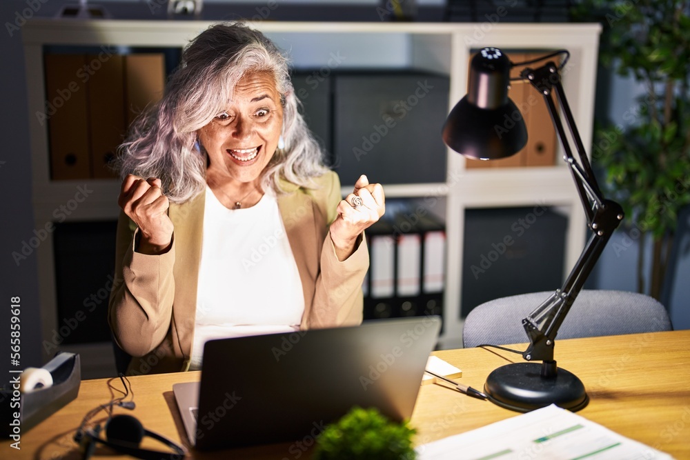 Middle age woman with grey hair working using computer laptop late at night celebrating ...