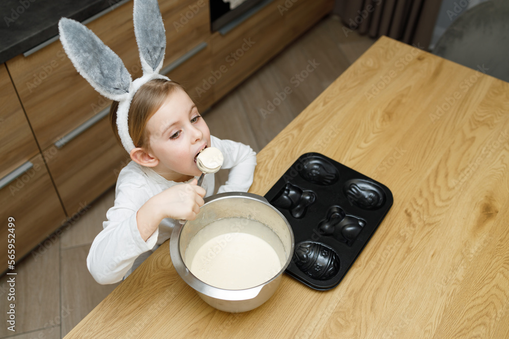 Little girl in bunny ears eating, licking sweet dough for muffins or ...