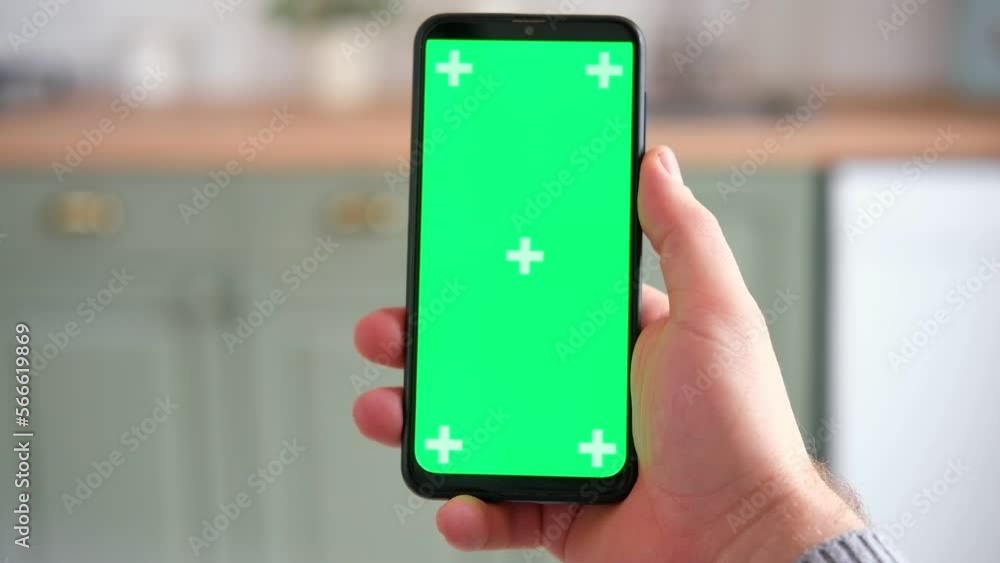 Young man sitting at home holding smartphone green mock-up screen in hand. Male person using ...