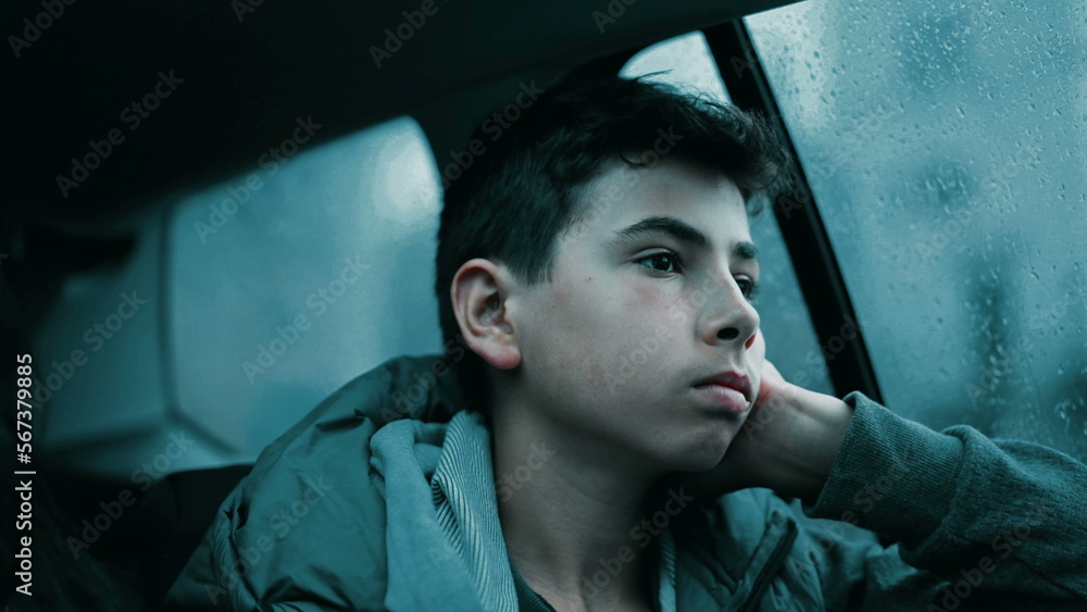 Depressed teen boy seated in car backseat looking out window during rainy day. Sad kid feeling ...