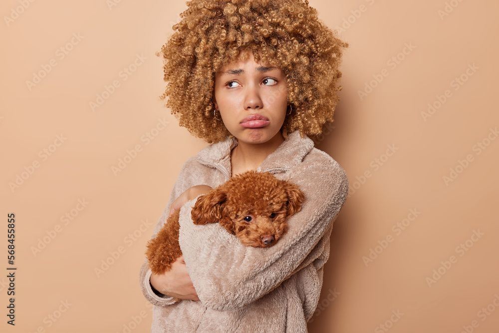 Unhappy curly haired young woman looks sadly aside embraces small ...