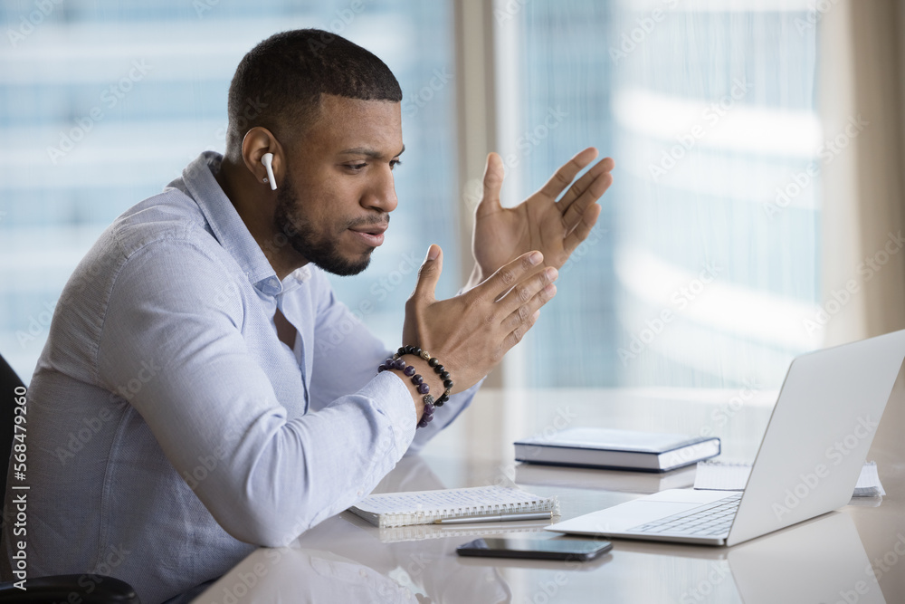 Motivated young African employee with earphone in ear talking on video ...